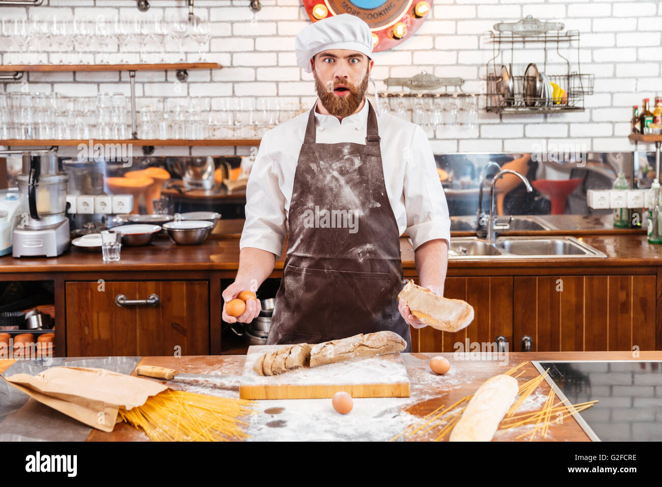 Surprised handsome baker cutting bread and holding eggs on the kitchen ...