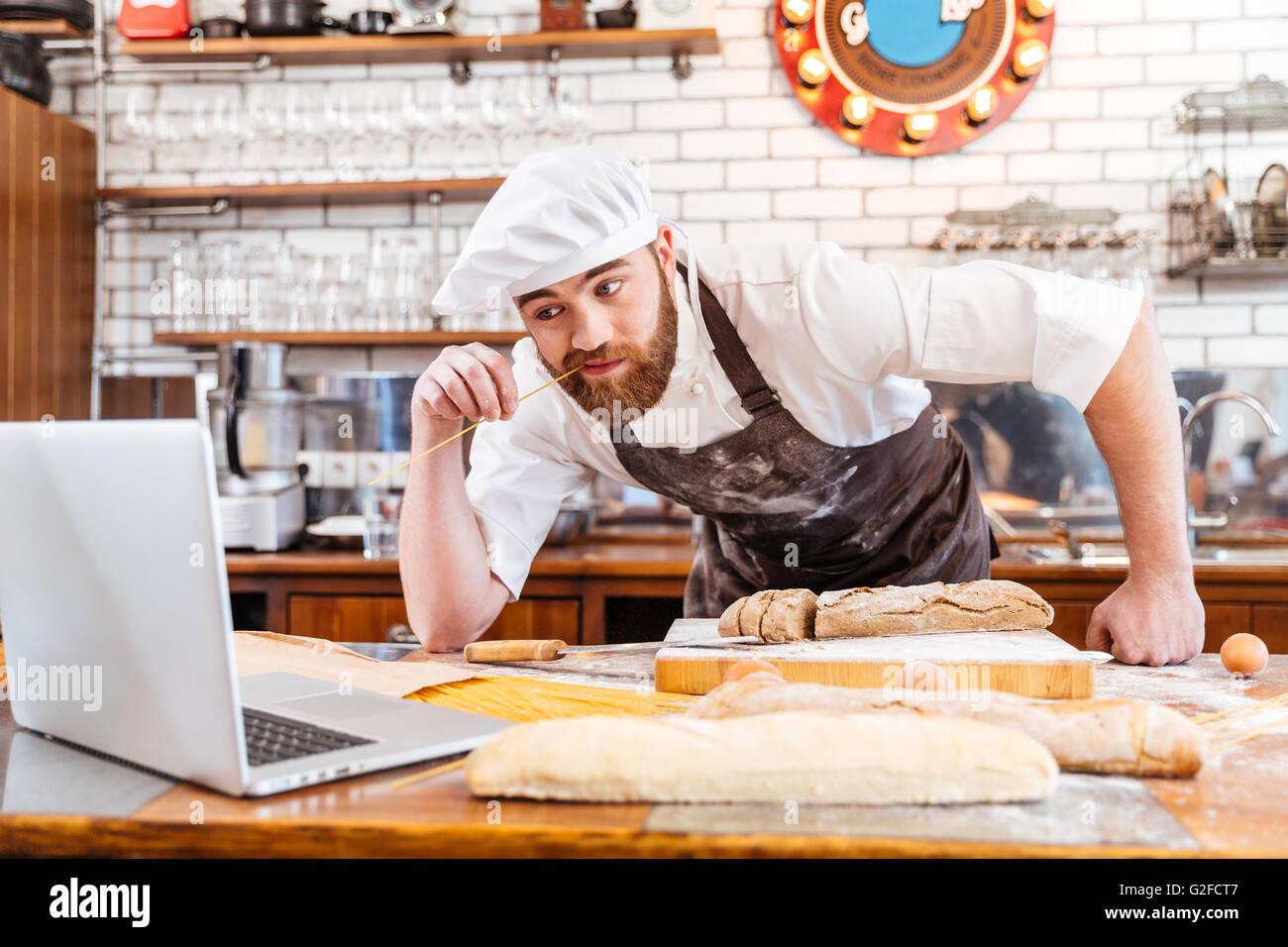 Thoughtful bearded baker cutting bread and using laptop on the kitchen ...