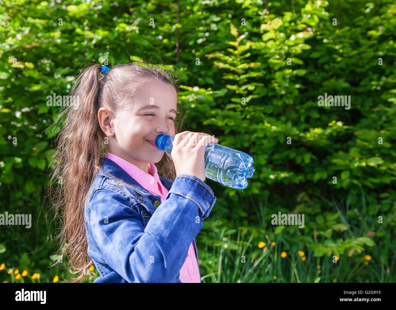 girl in a blue denim jacket, drinks water from a plastic bottle outside ...