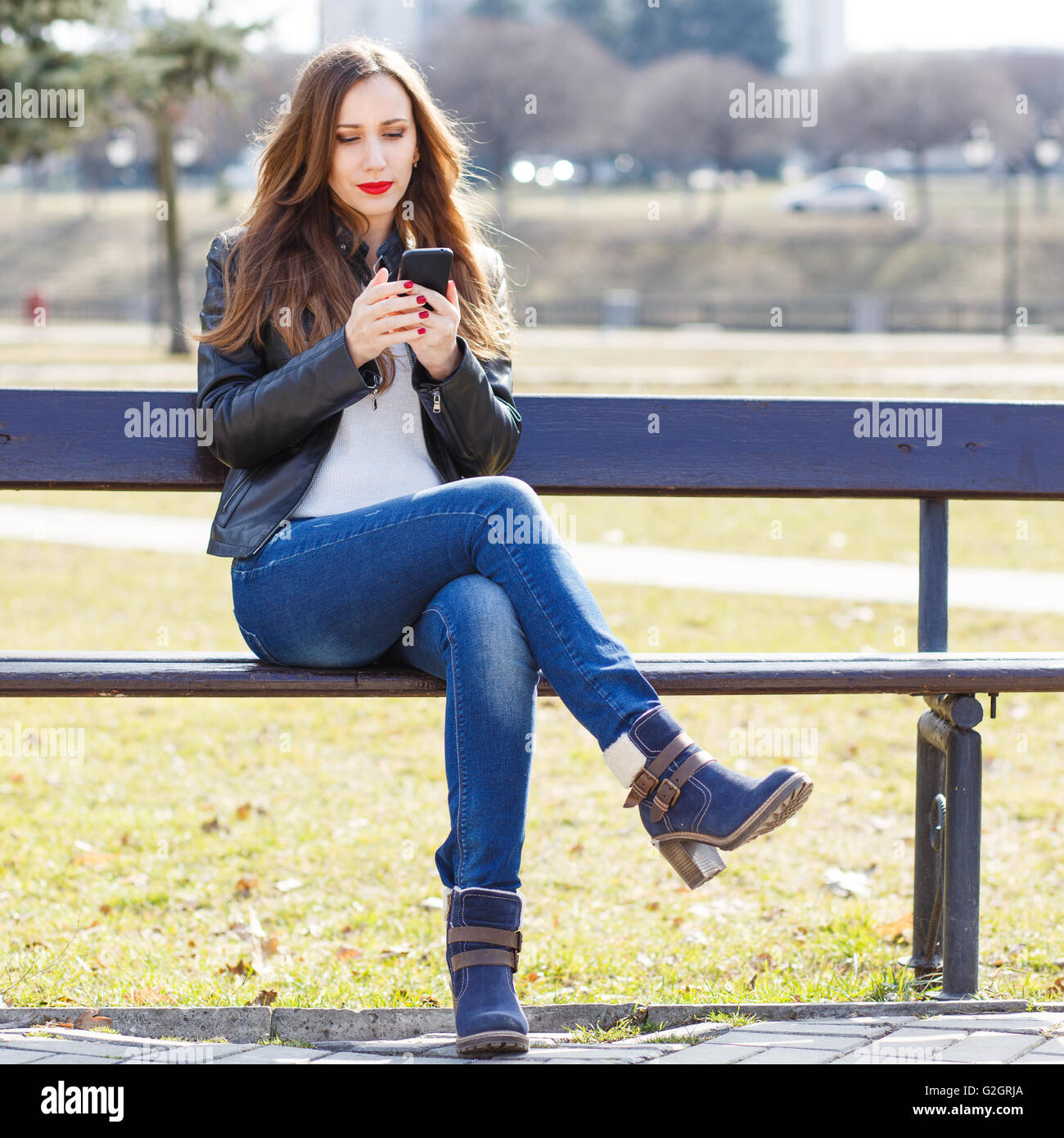 Young smiling woman using smartphone sitting on bench in park. Beautiful european girl texting ...