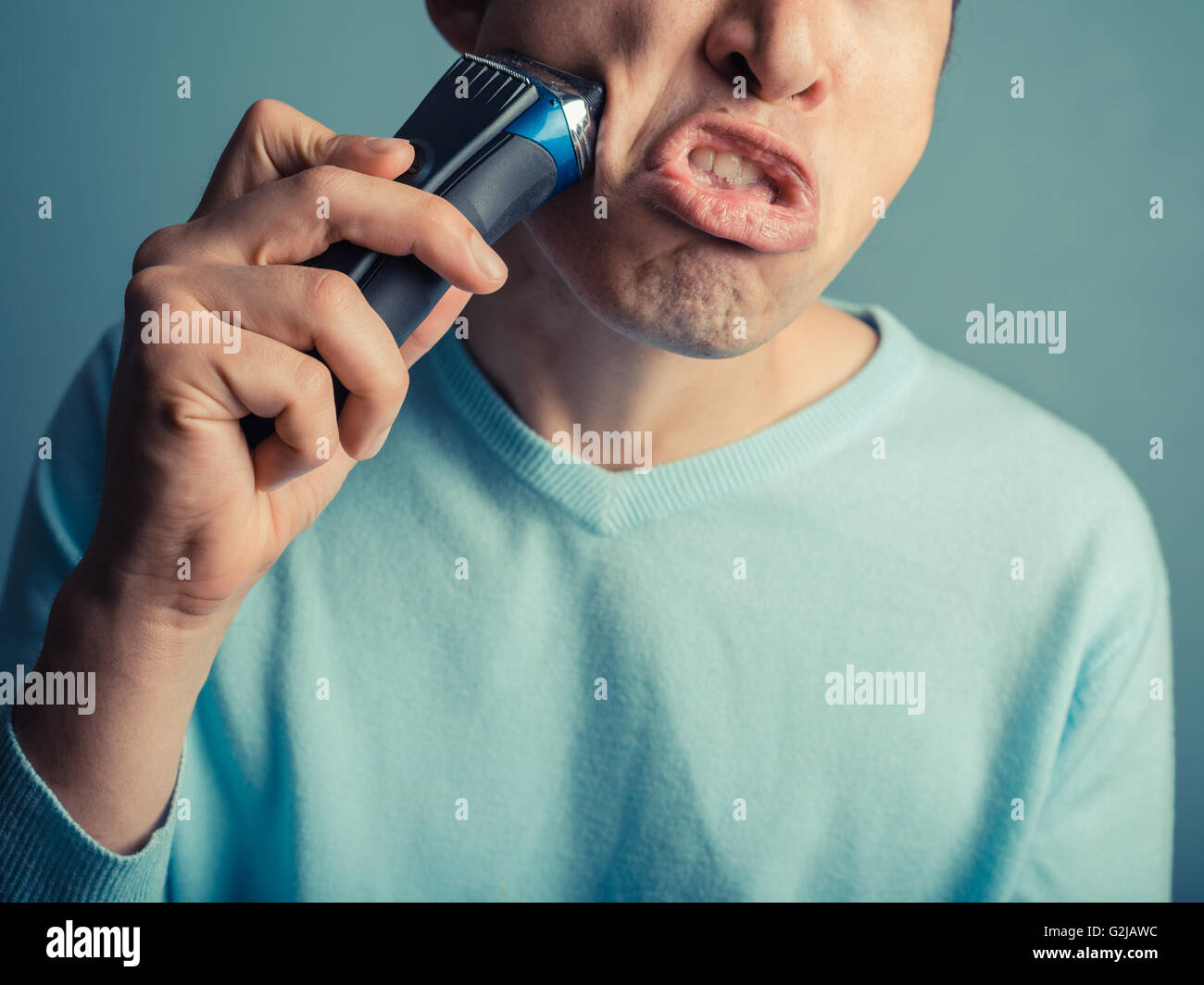 A young man is shaving with an electric razor and is pulling faces ...