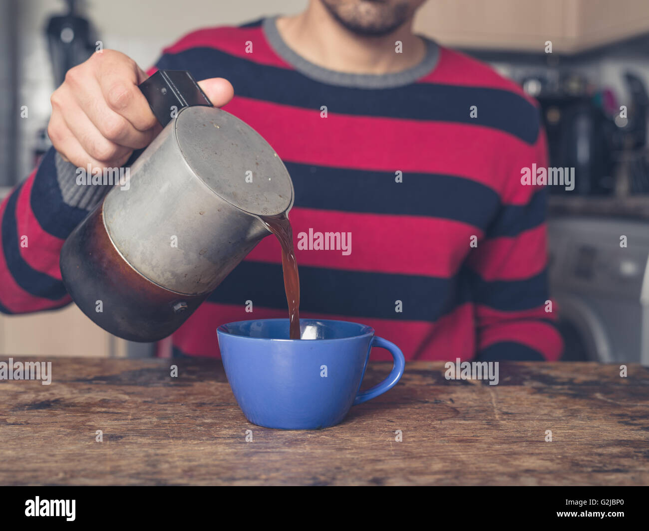 A young man is sitting at a table in a kitchen and is pouring coffee ...