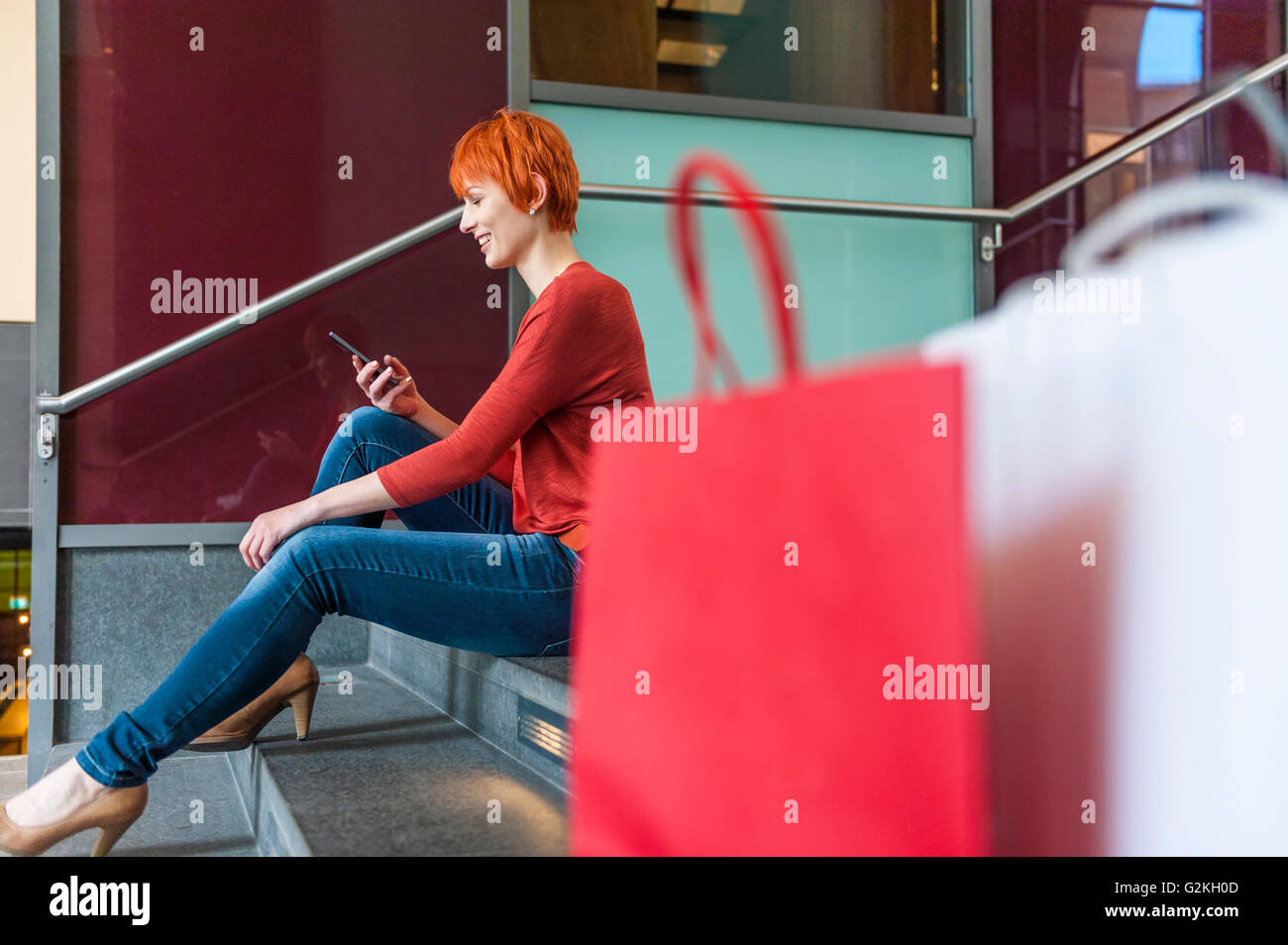 Young woman sitting on stairs with cell phone and shopping bags next to ...