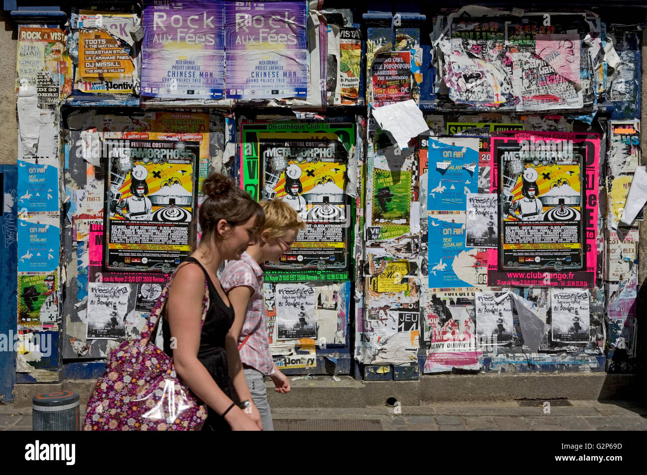 Two girls walk past rock posters and flyers covering a wall in Rue Pont ...