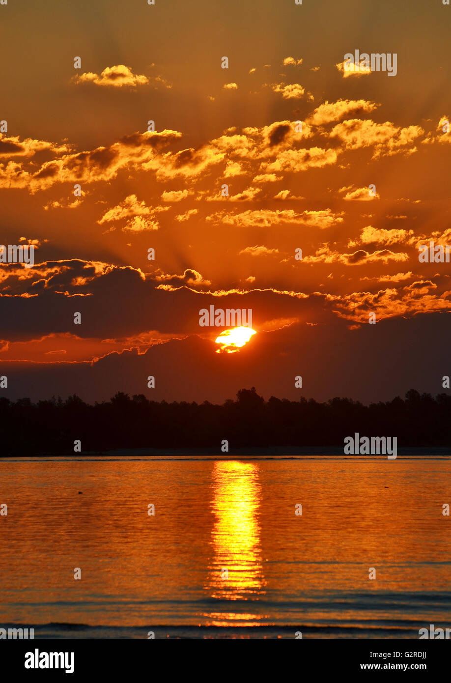 Glowing orange sun setting (sunset) behind clouds with reflections on ...