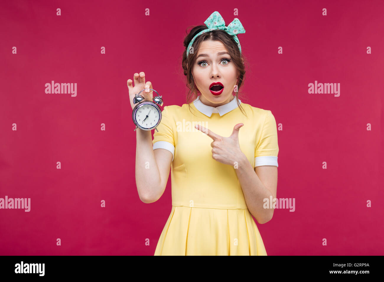 Cute amazed pinup girl pointing on alarm clock over pink background ...