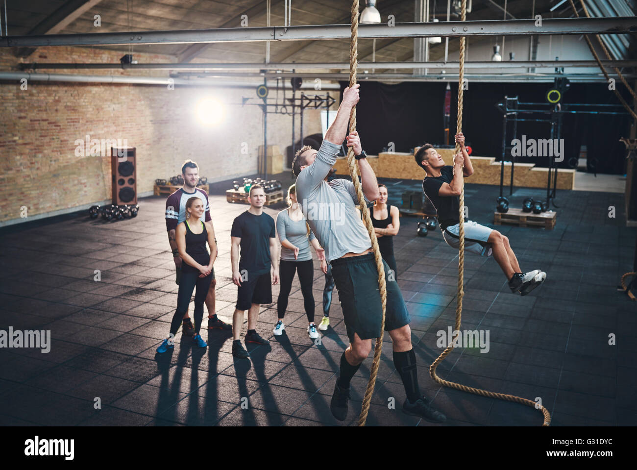 Two fit young men climbing ropes in a gym with people on the floor ...