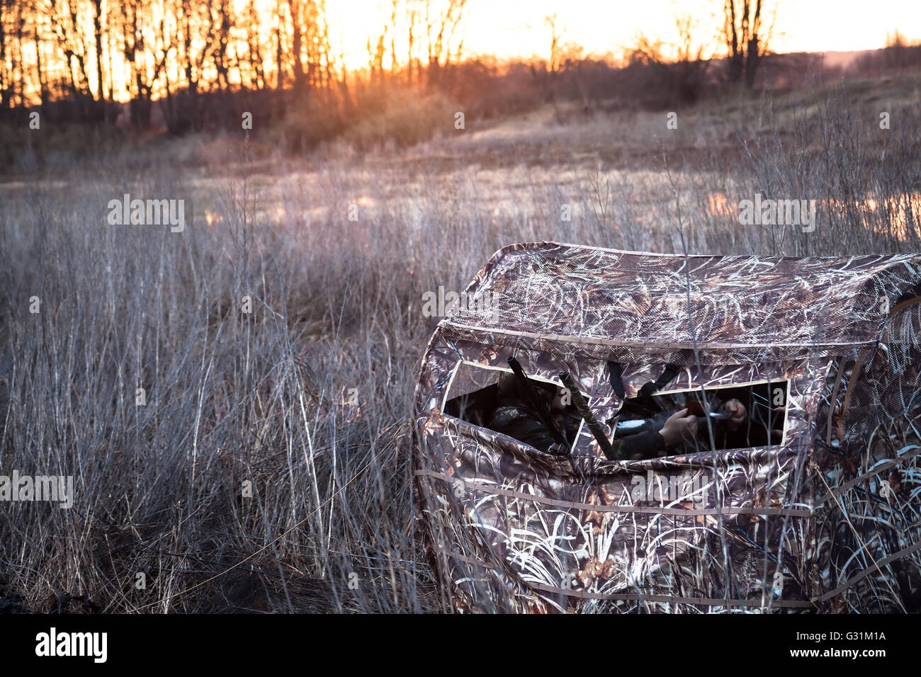 Hunting tent with hunters waiting for prey in the field next to the river while picturesque ...