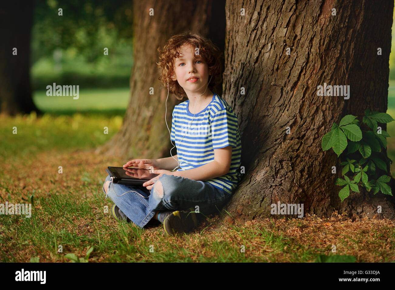 The boy of 8-9 years sits in park under a tree with the tablet in hands ...