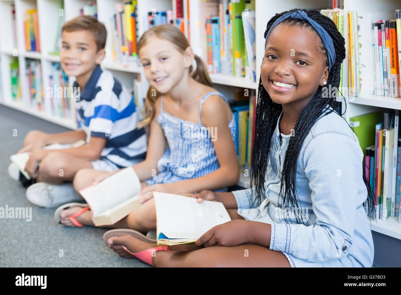 Portrait of school kids sitting on floor and reading book in library ...