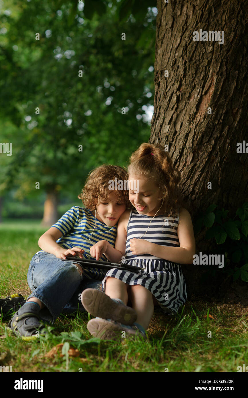 Two children sit in park under a big tree and play on the tablet ...