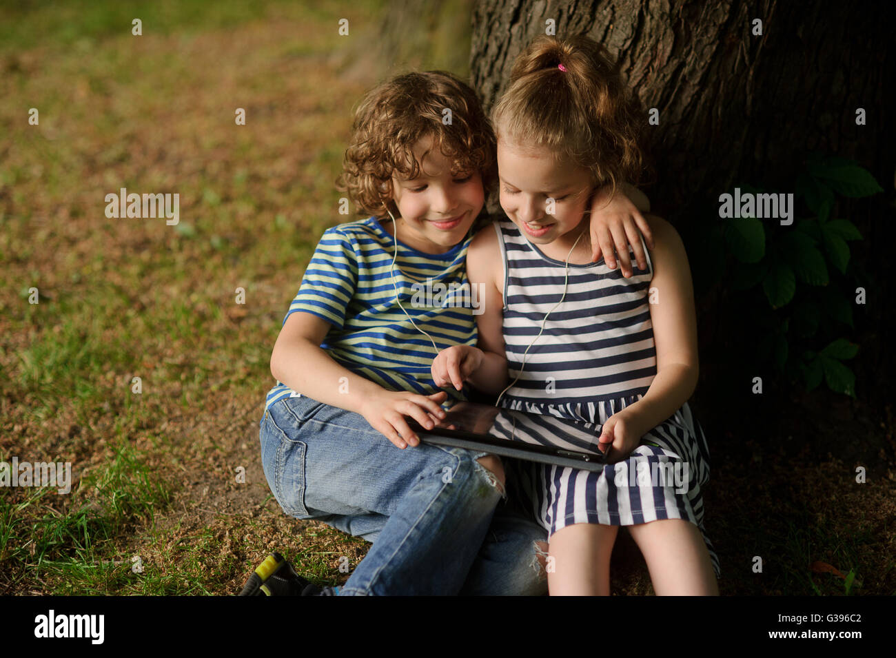 Two children sit under more tree with interest looking at the tablet ...