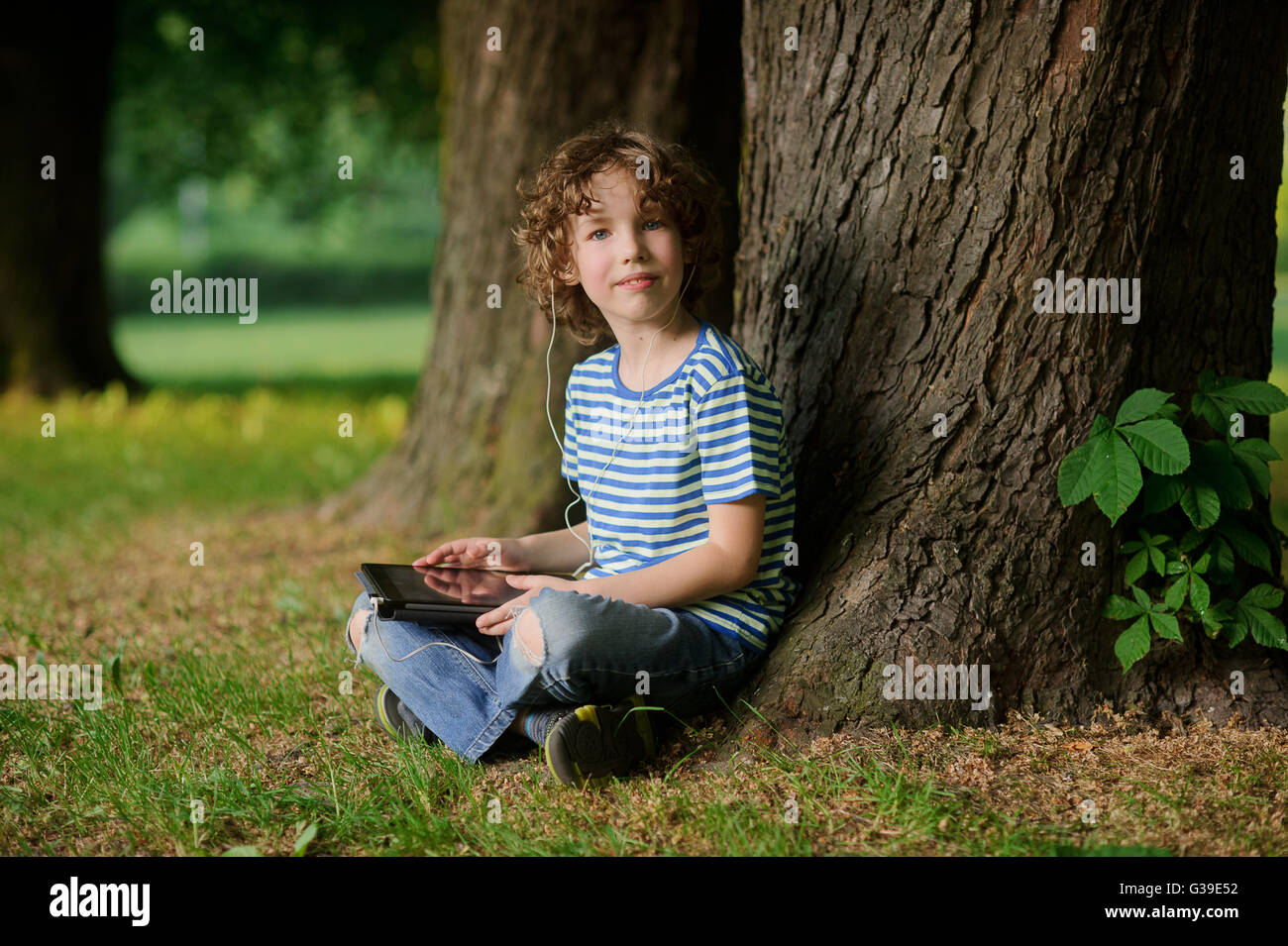 The boy of 8-9 years sits leaning against a tree and holds the tablet ...
