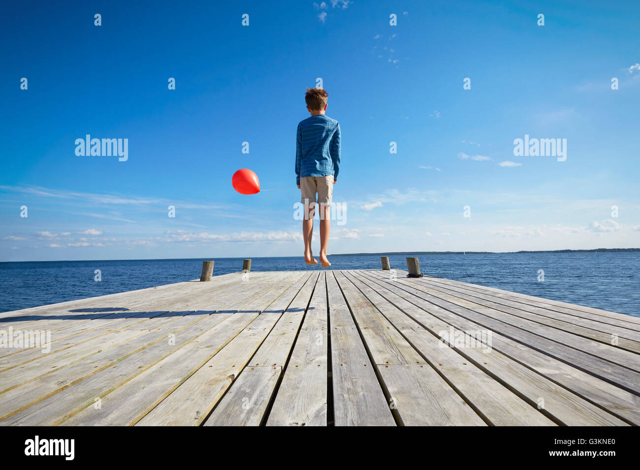 Young boy jumping on wooden pier, holding red helium balloon, rear view ...