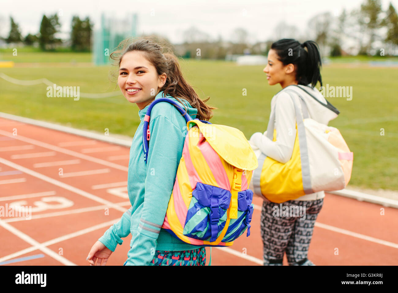 Two young women walking on running track, carrying sports bags, rear ...