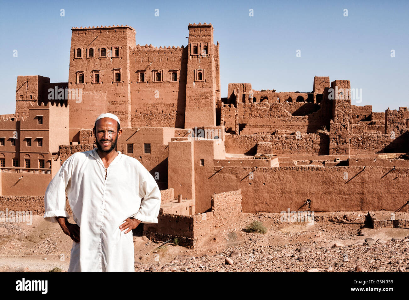 Moroccan man in traditional long white clothing stands near kasbah fort ...