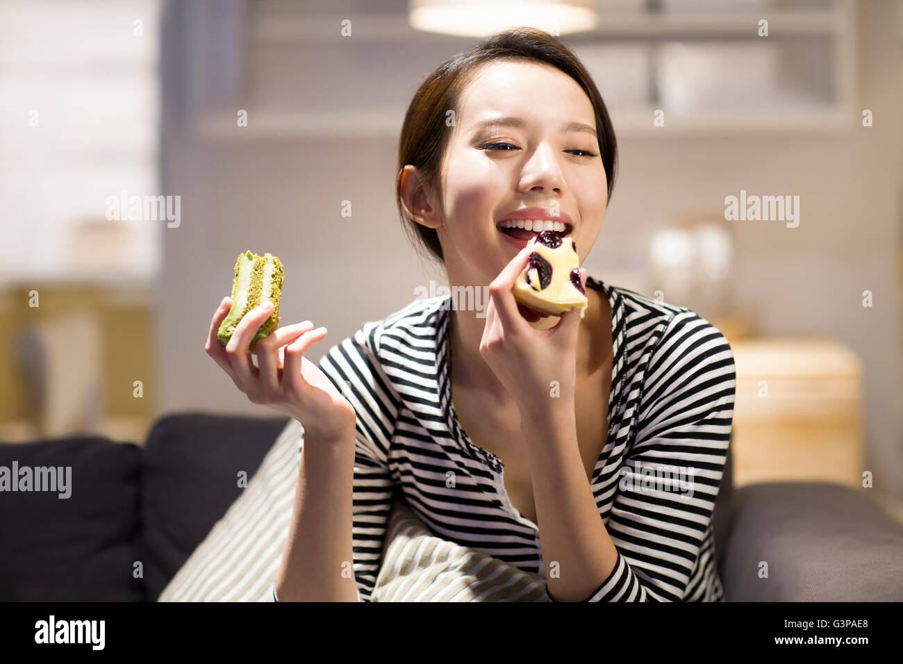 Happy young woman eating cakes on sofa Stock Photo - Alamy
