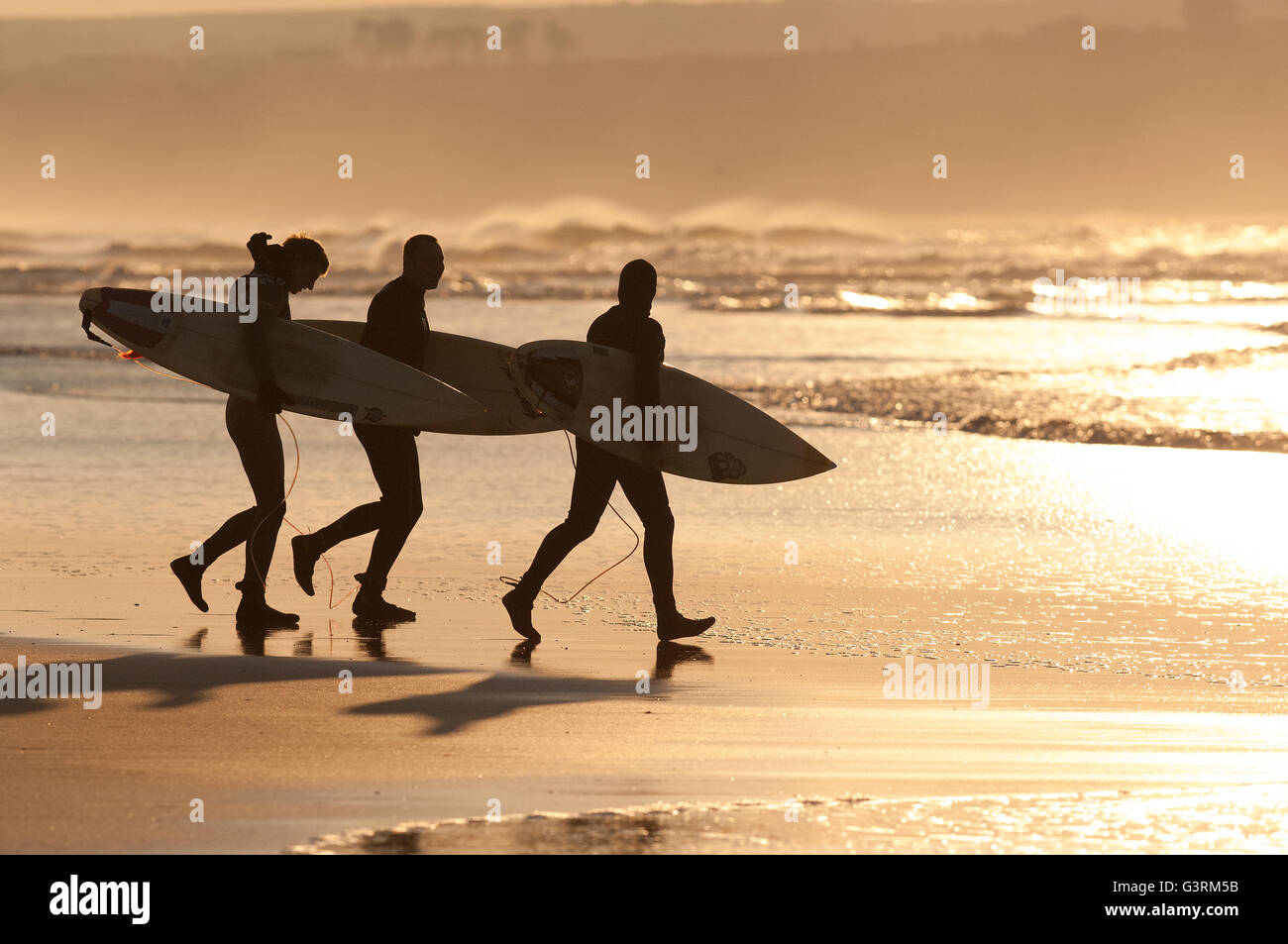 Three Surfers carrying surf boards on the beach in silhouette at sunset ...