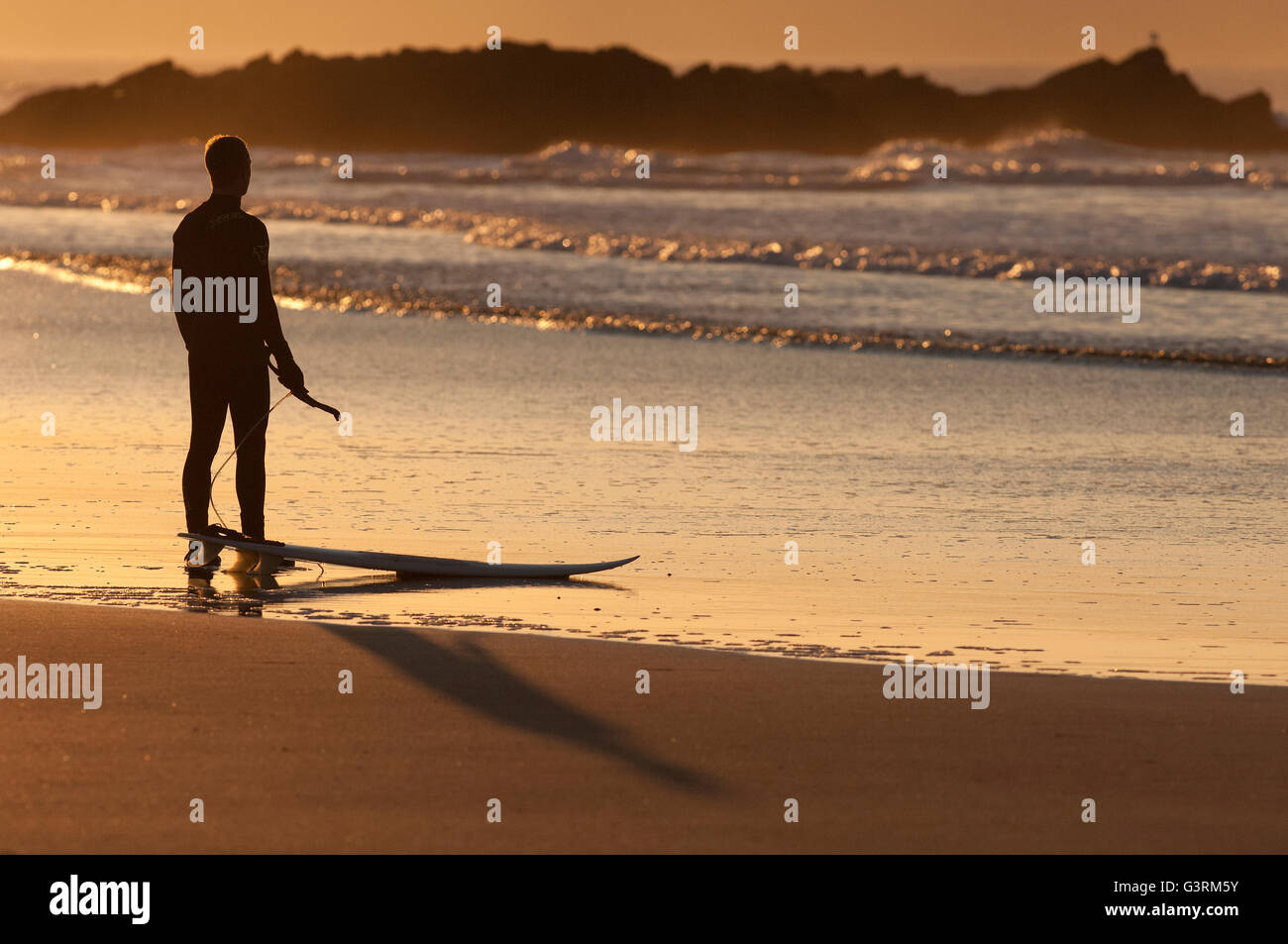 Surfer in silhouette casting long shadow on the sand watching the surf ...