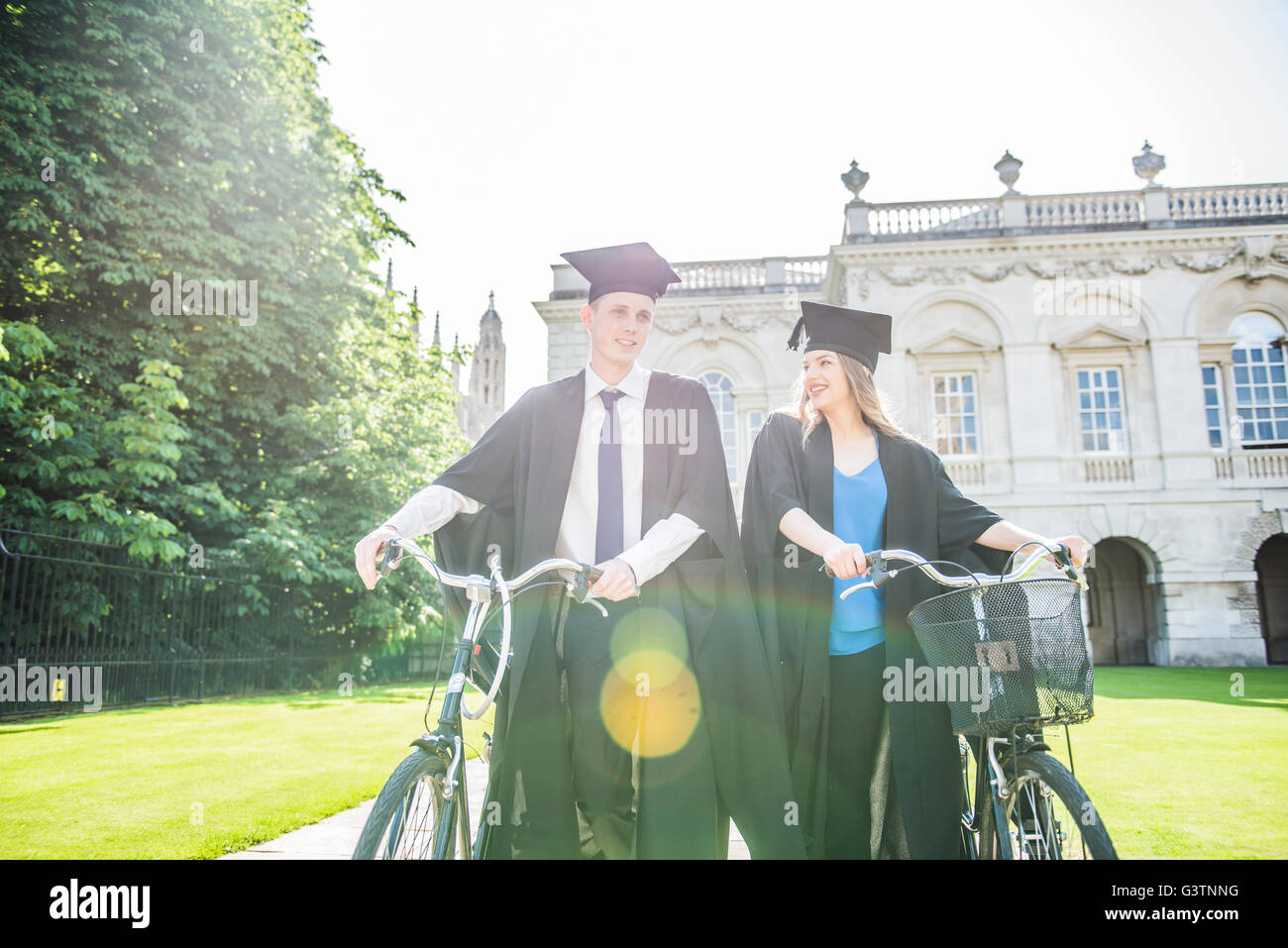 Two young students in graduation gowns pushing bicycles through the ...