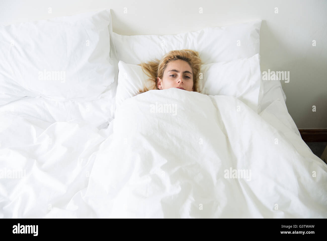 A cool young woman lying in a bed with he covers pulled up to her chin ...