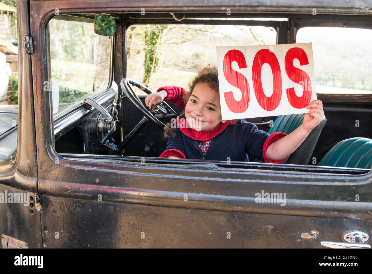 A young girl holding an SOS sign up in the window of a vintage car ...