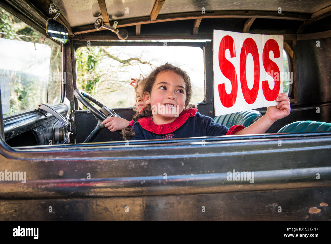 A young girl holding an SOS sign up in the window of a vintage car ...