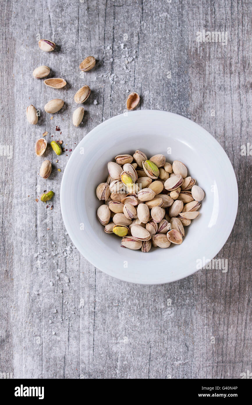 Salted roasted pistachios nuts with shell and salt in white ceramic plate over white wooden ...