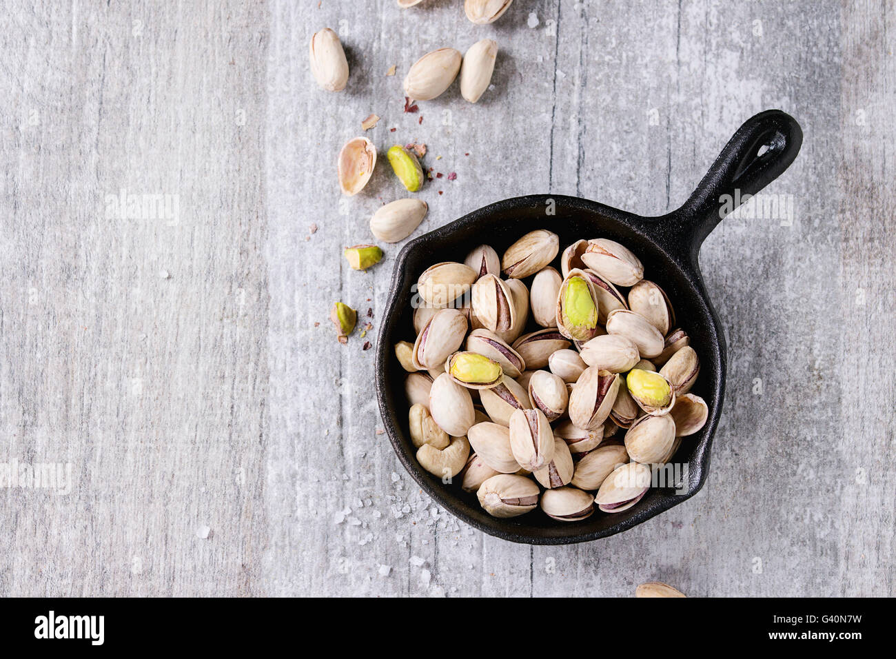 Salted roasted pistachios nuts with shell and salt in black small cast-iron pan over white ...
