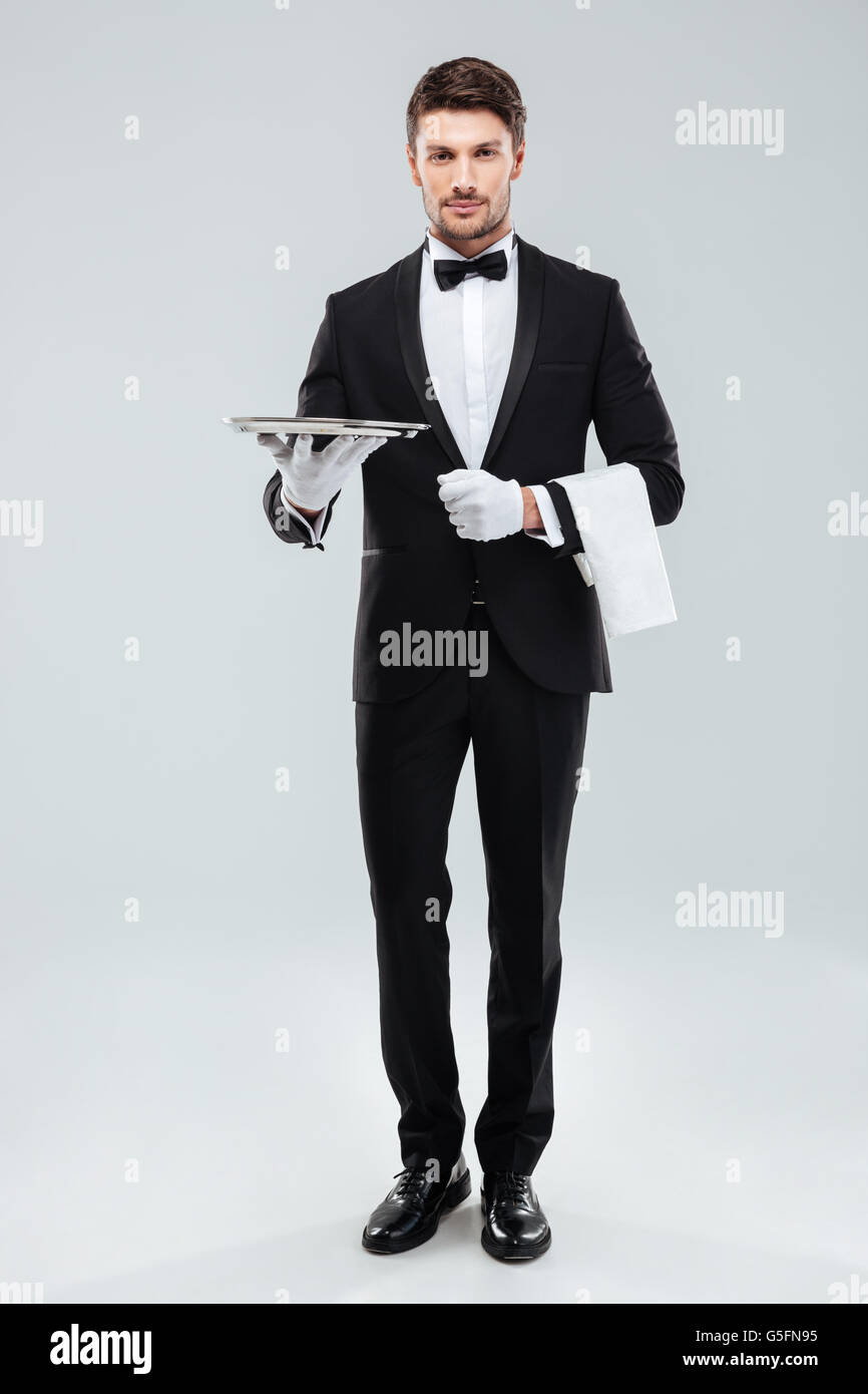 Full length of confident young waiter in tuxedo standing and holding ...