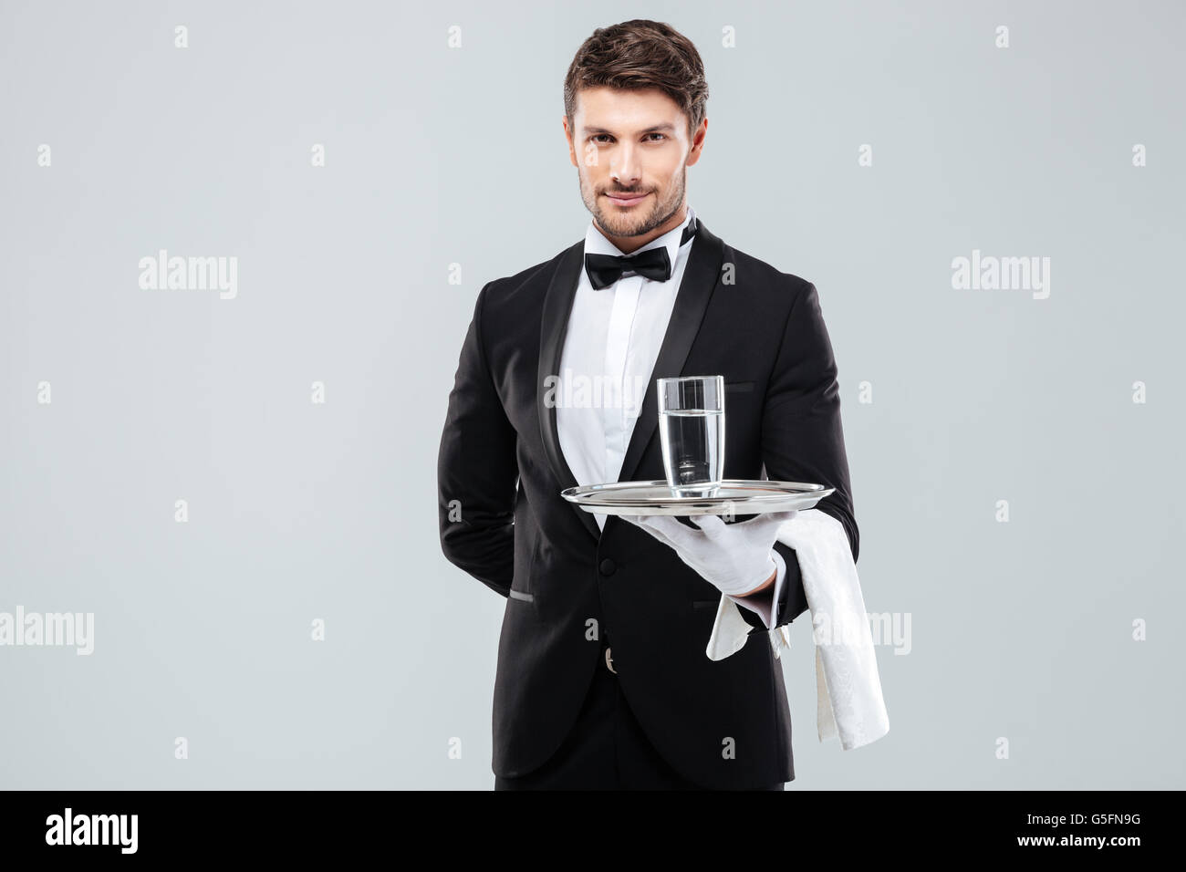 Confident young butler in tuxedo and gloves holding glass of water on ...