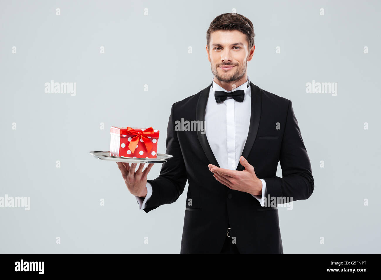 Smiling young waiter in tuxedo standing and holding present box on tray ...