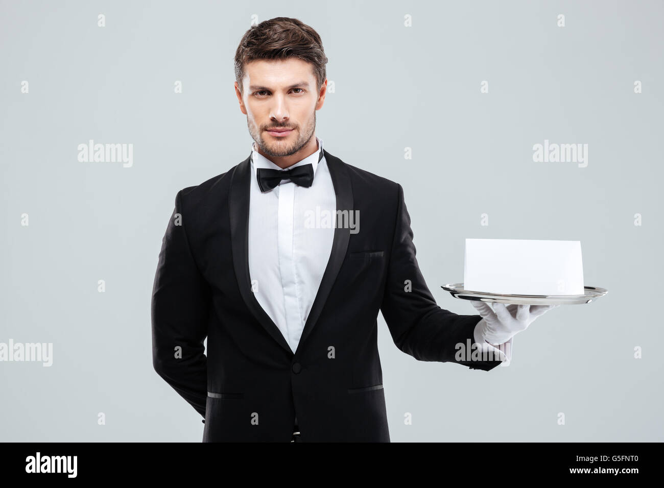 Attractive young butler in tuxedo and gloves holding tray with blank ...