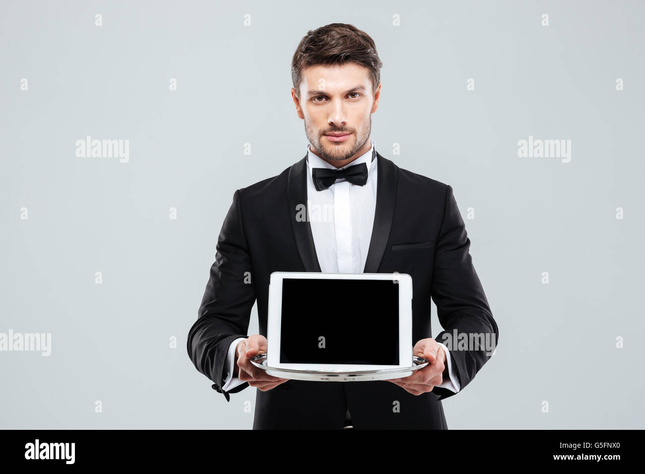 Handsome young butler in tuxedo with bowtie holding blank screen tablet ...