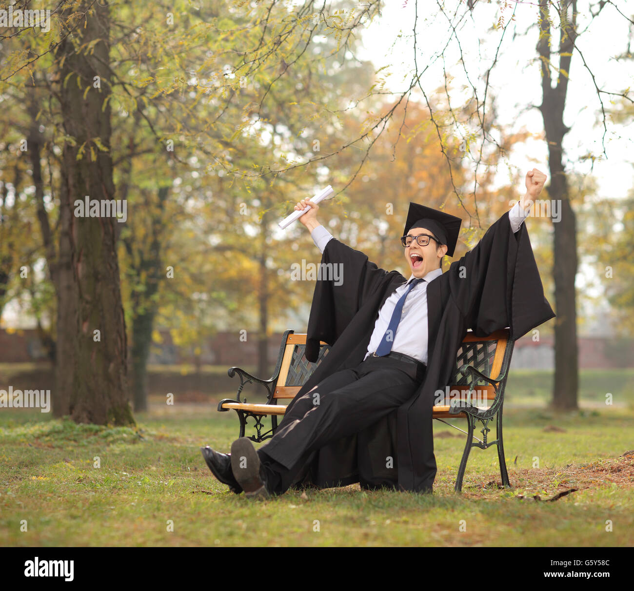Joyful student celebrating his graduation seated on a bench in a park ...
