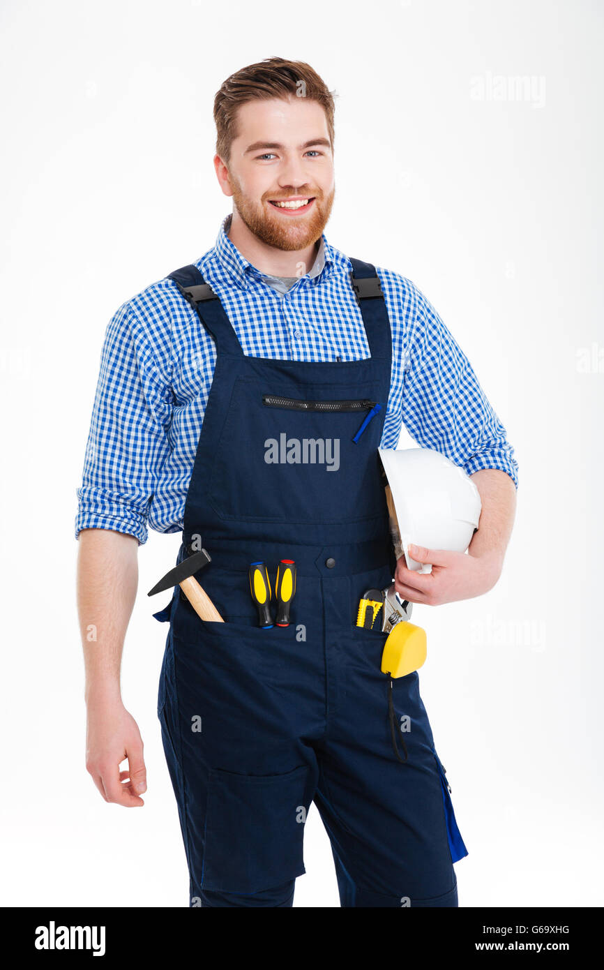 Cheerful handsome young builder in overall standing and holding helmet ...