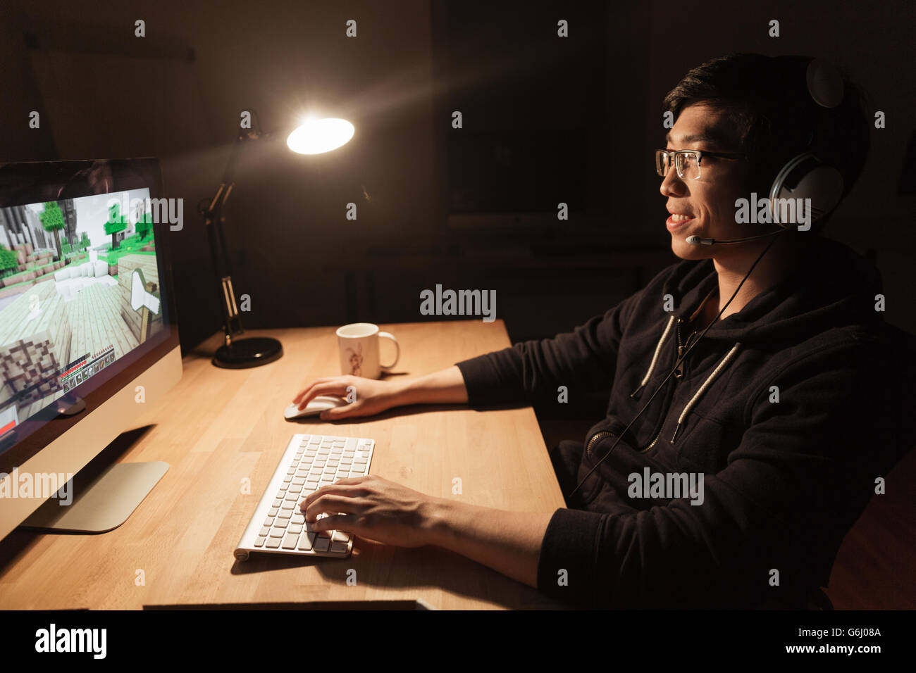 Smiling asian young male gamer playing computer game in dark office ...
