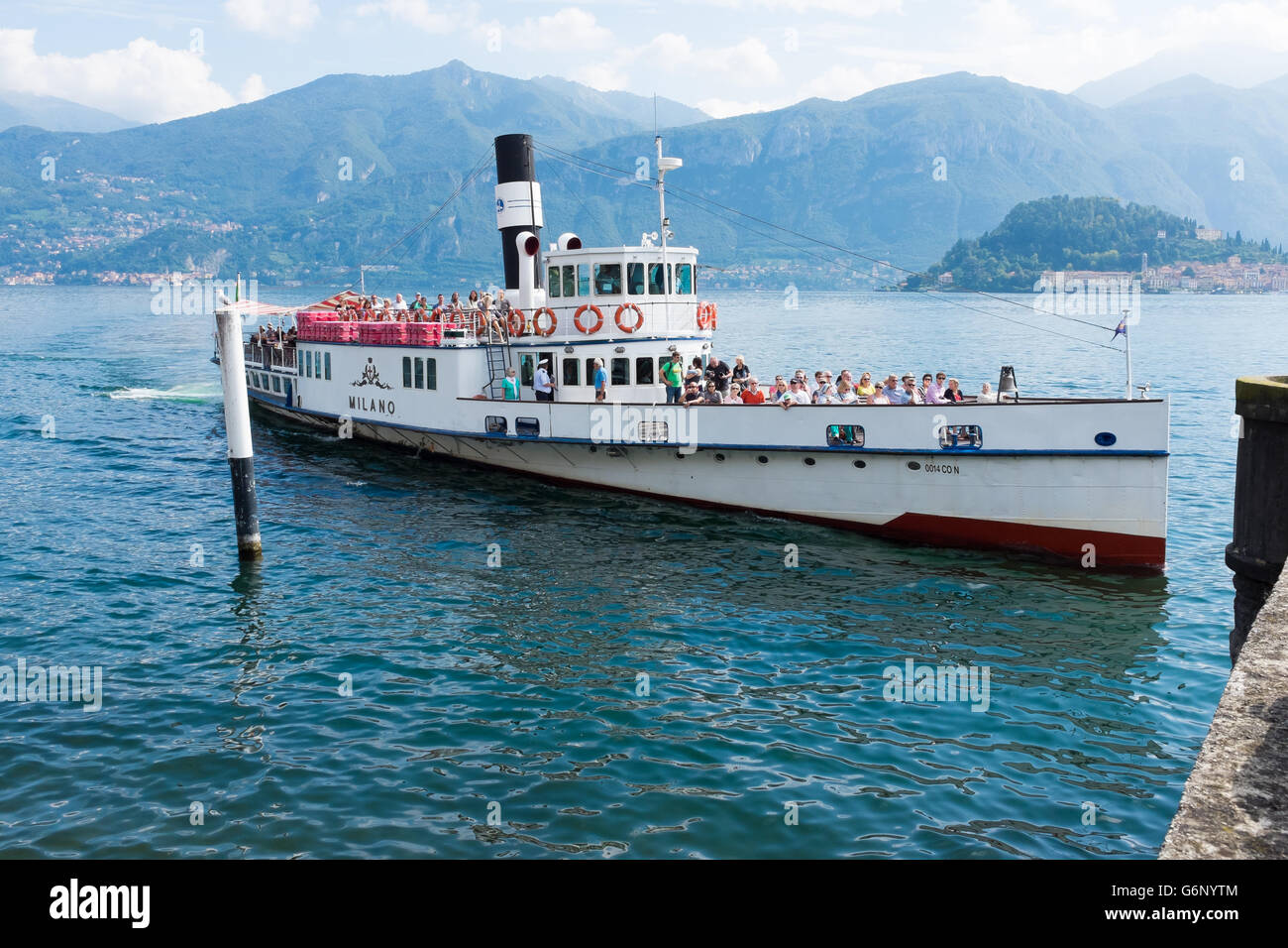 A ferry nears the dock on the shore of Lake Como. There are mountains ...