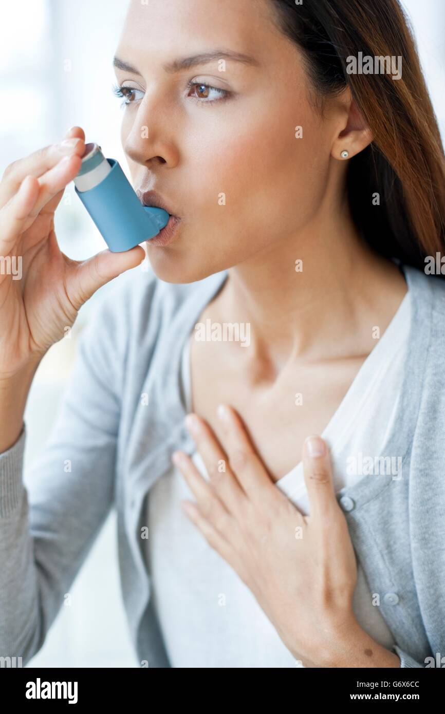 MODEL RELEASED. Young woman using an inhaler with her hand on her chest ...