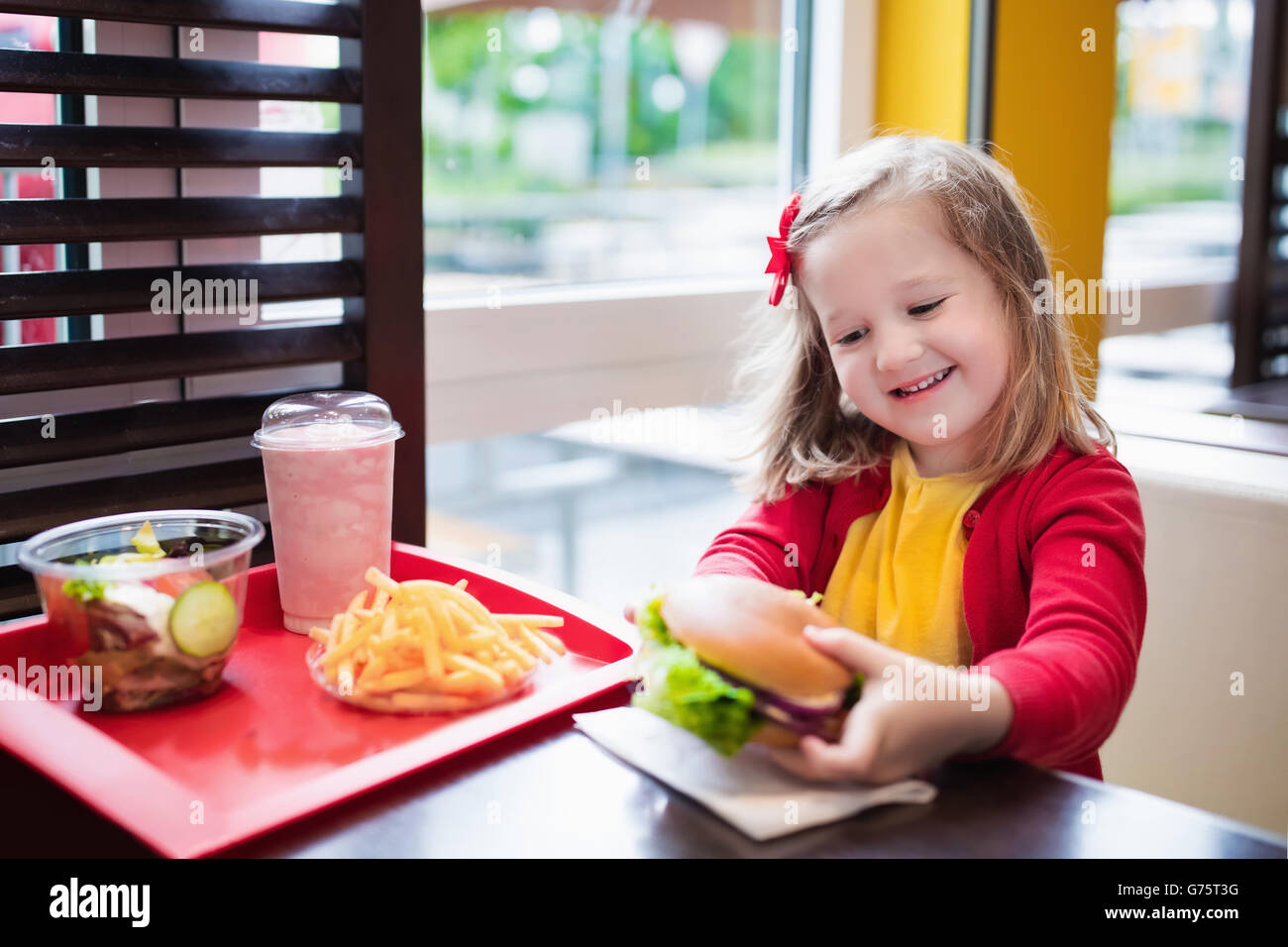 Little girl eating hamburger and French fries in a fast food restaurant ...