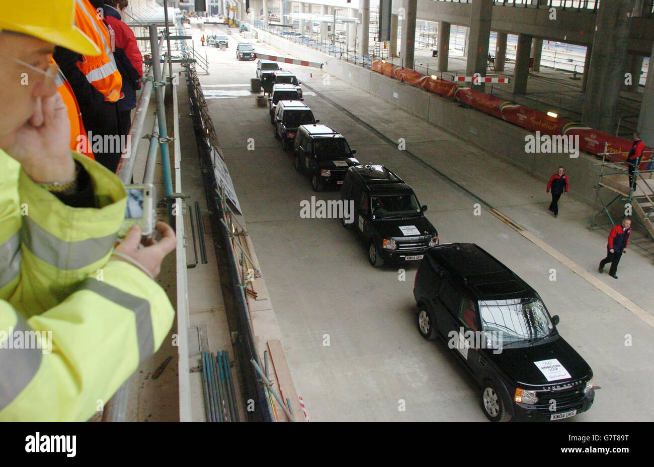 Contractors watch as delegates from the IOC are driven by Land Rover ...