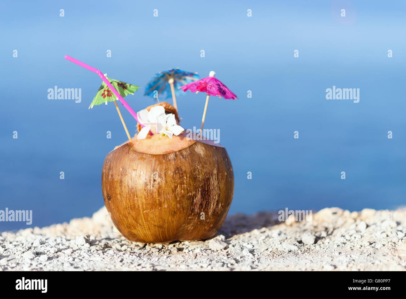 Coconut with drinking straw, umbrellas and flowers, on rocks at the sea ...