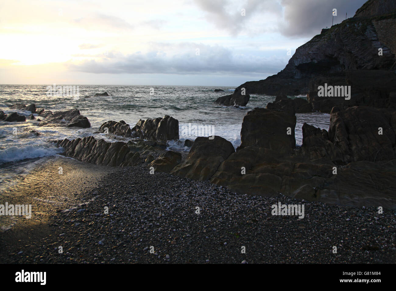 A sombre evening shore with silhouetted jagged rocks and pebble beach ...