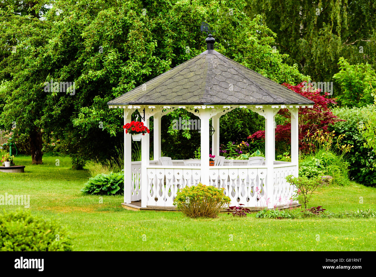 Lovely white gazebo with red flowers hanging from a basket. Trees and ...