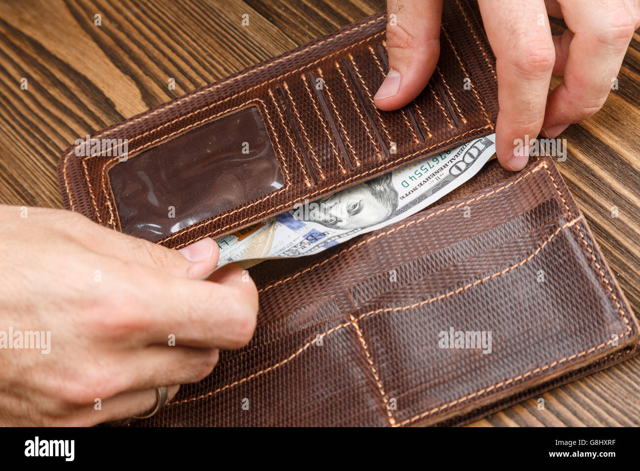 man is putting 100 dollar banknote in a brown leather wallet over dark ...
