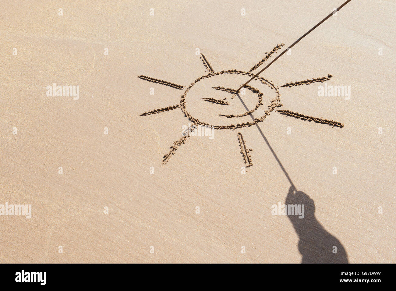 Shadow hand holding a stick drawing a smiley face sun on a beach. UK ...