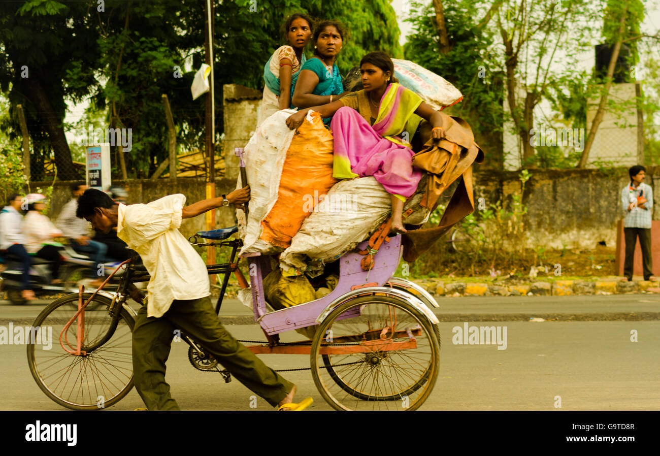 A Rickshaw puller tugs his rickshaw with a load of stuff & three ladies ...