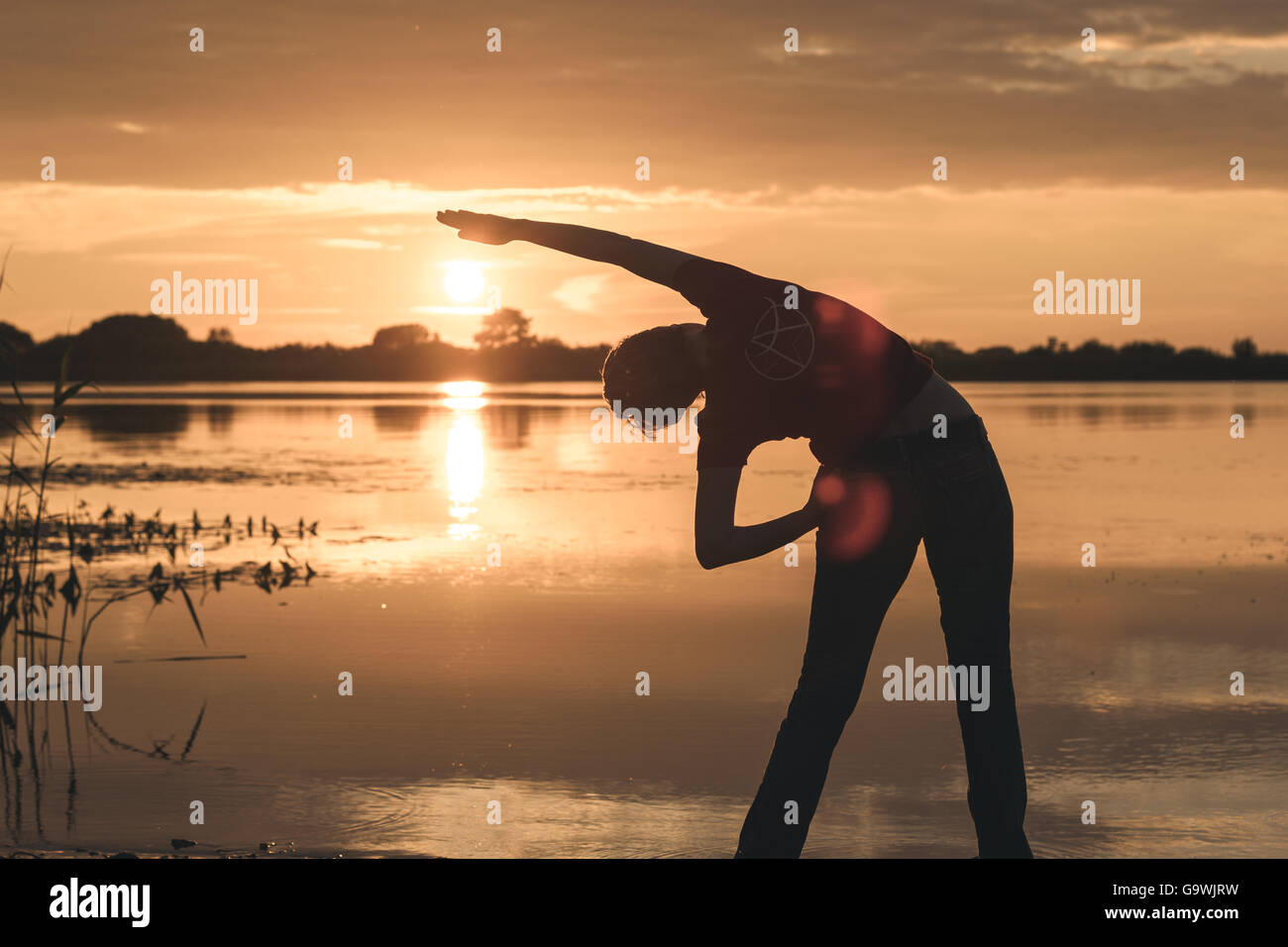 young slim woman doing exercises on the shore of the lake at sunset ...