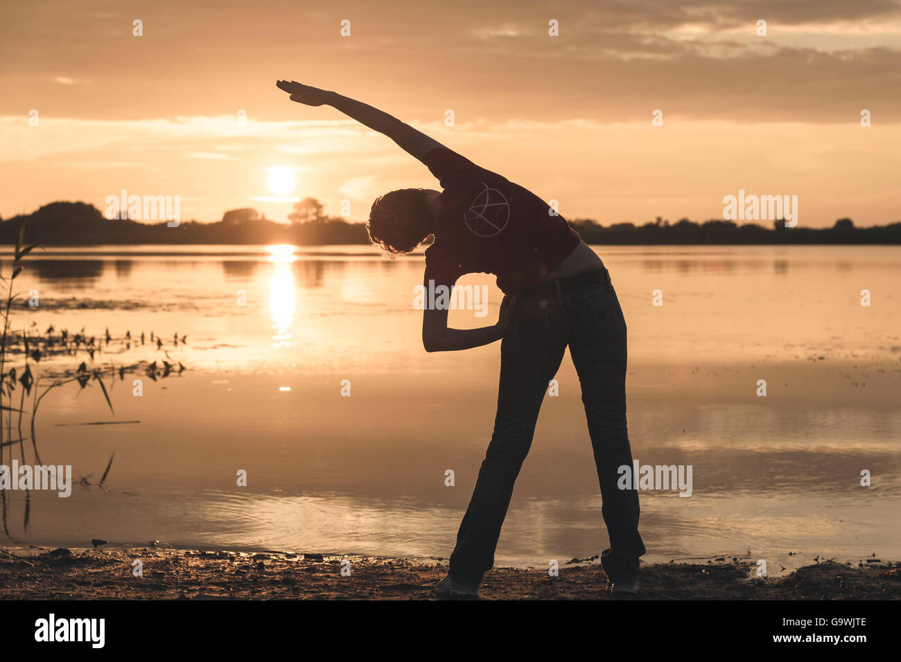 young slim woman doing exercises on the shore of the lake at sunset ...