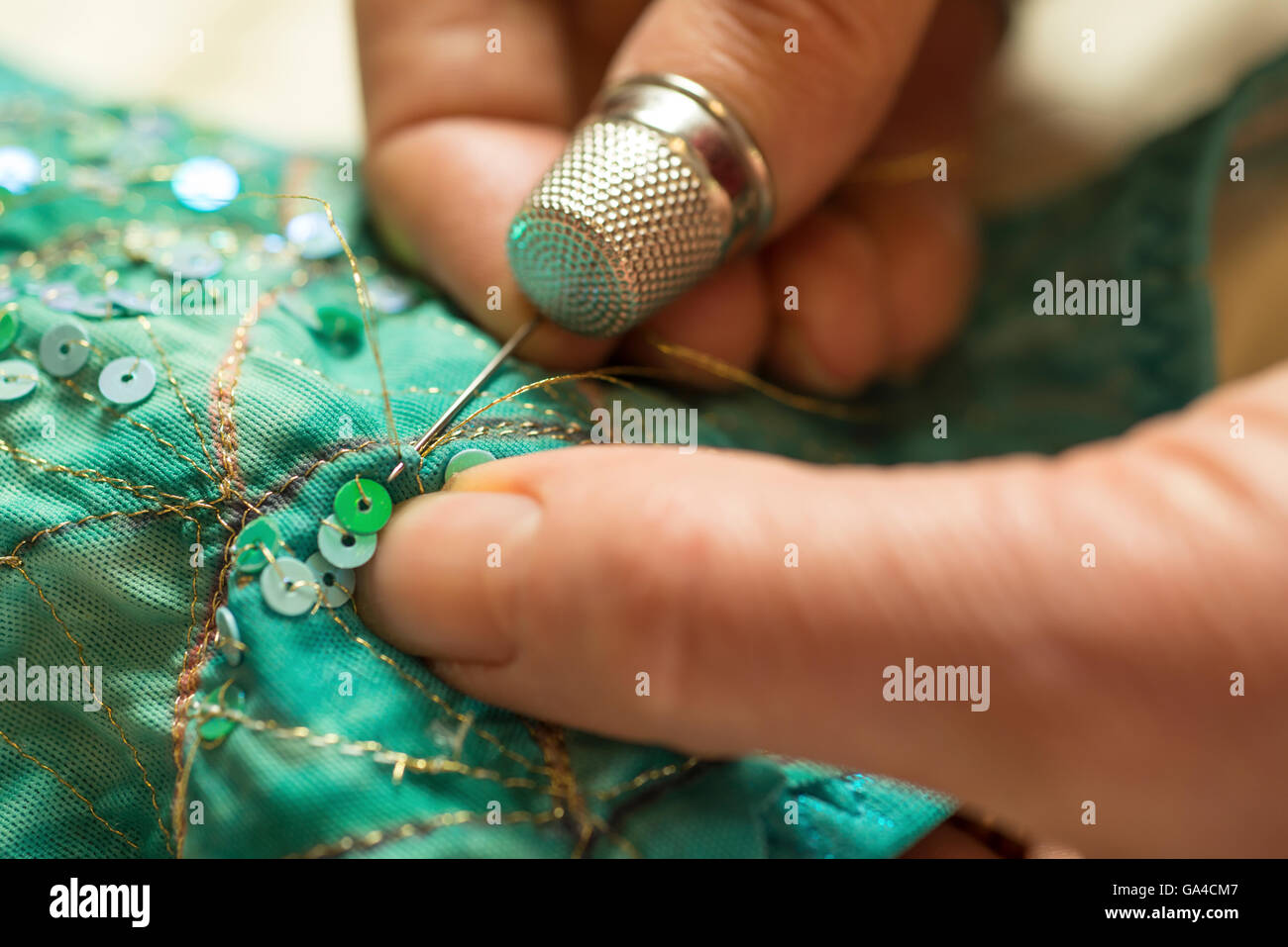 Hand with thimbled thumb pushing needle with gold thread through green ...